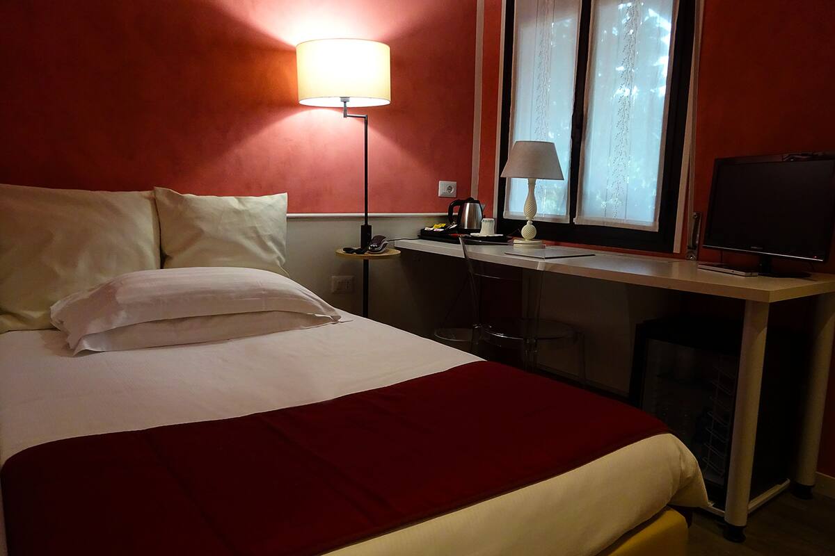 Quarto de hotel com cama, escrivaninha, televisão e janela com cortinas.