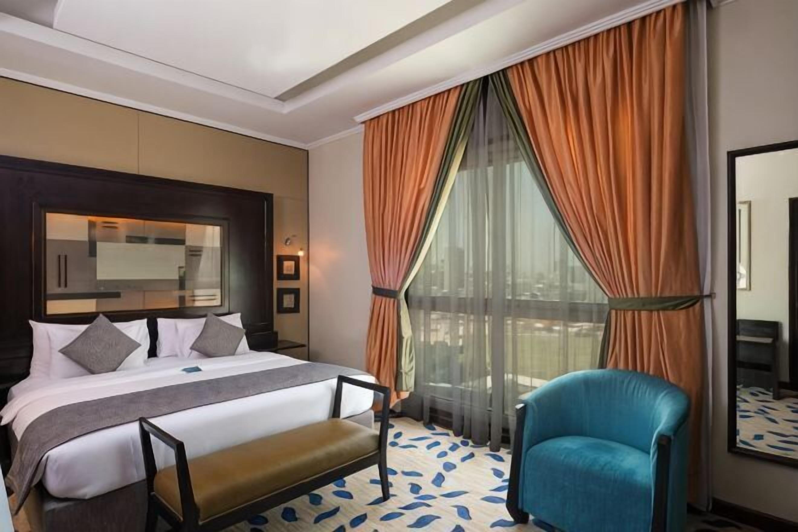 Quarto de hotel com uma cama grande, um banco, uma poltrona azul e vista para a cidade.