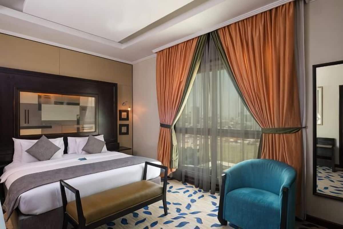 Quarto de hotel com uma cama grande, um banco, uma poltrona azul e vista para a cidade.