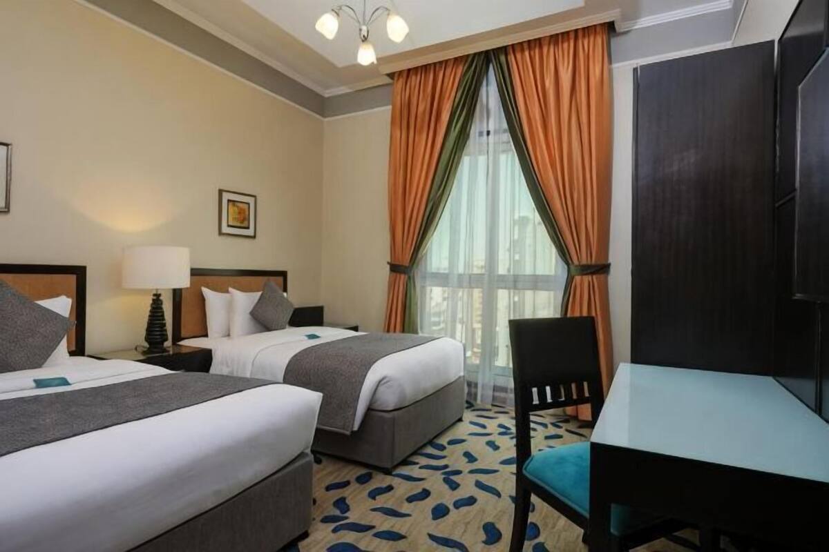 Quarto de hotel com duas camas, uma escrivaninha, uma cadeira, televisão e janela com cortinas.