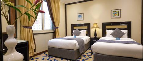 Quarto de hotel com duas camas, mesa de cabeceira com luminária, vaso com flores e janela com cortinas.