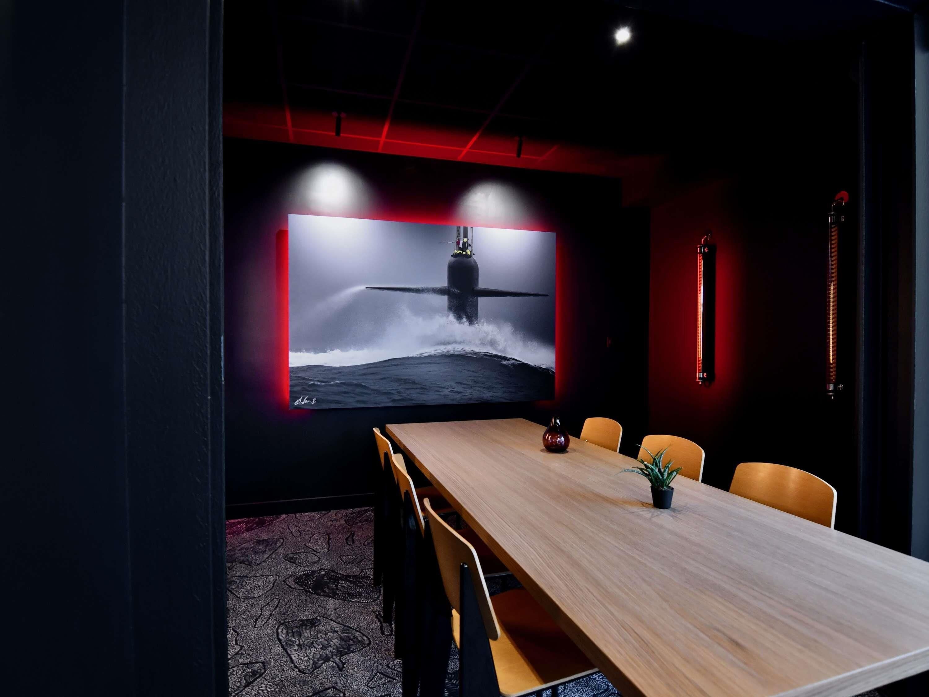 Sala de conferência com uma grande tela fixada na parede exibindo um submarino, uma mesa de madeira e quatro cadeiras.