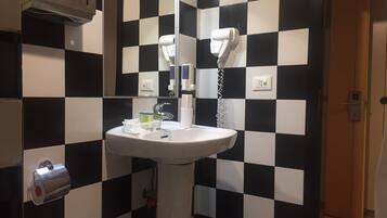 Um banheiro com parede de azulejos quadriculada em preto e branco, pia branca, dois secadores de cabelo fixados na parede e um secador de mãos no chão.