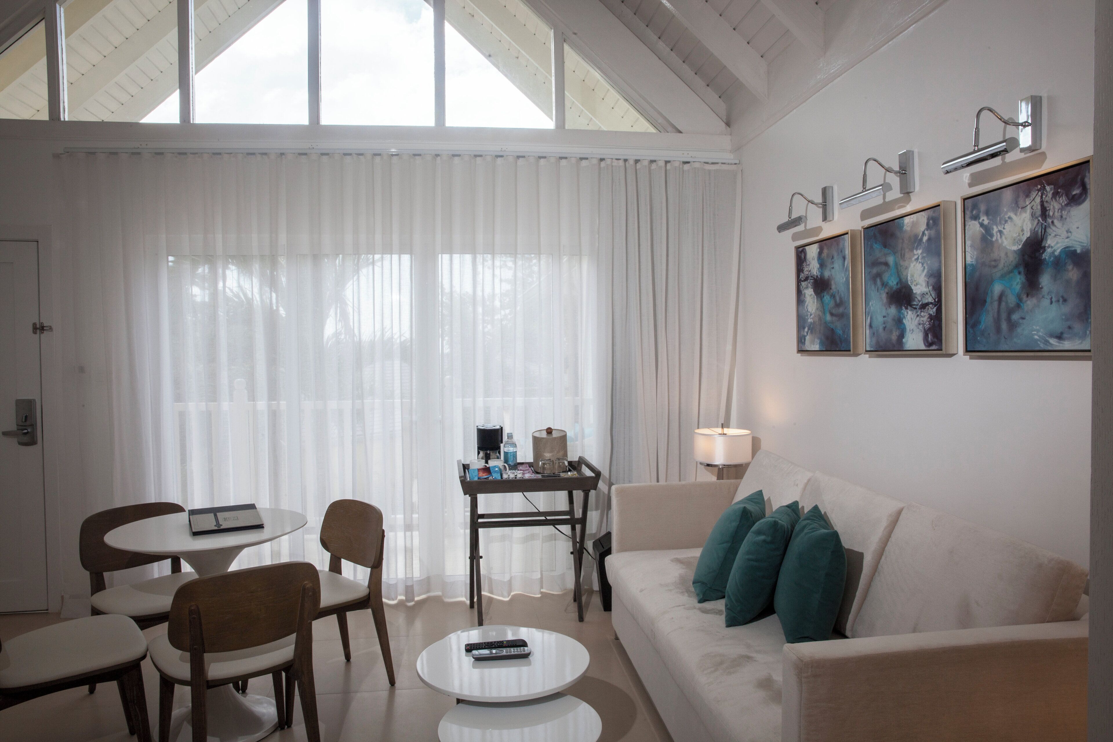 Uma sala de estar moderna com sofá branco, almofadas verde-turquesa, uma mesa de centro redonda e uma área de jantar com cadeiras e uma mesinha.