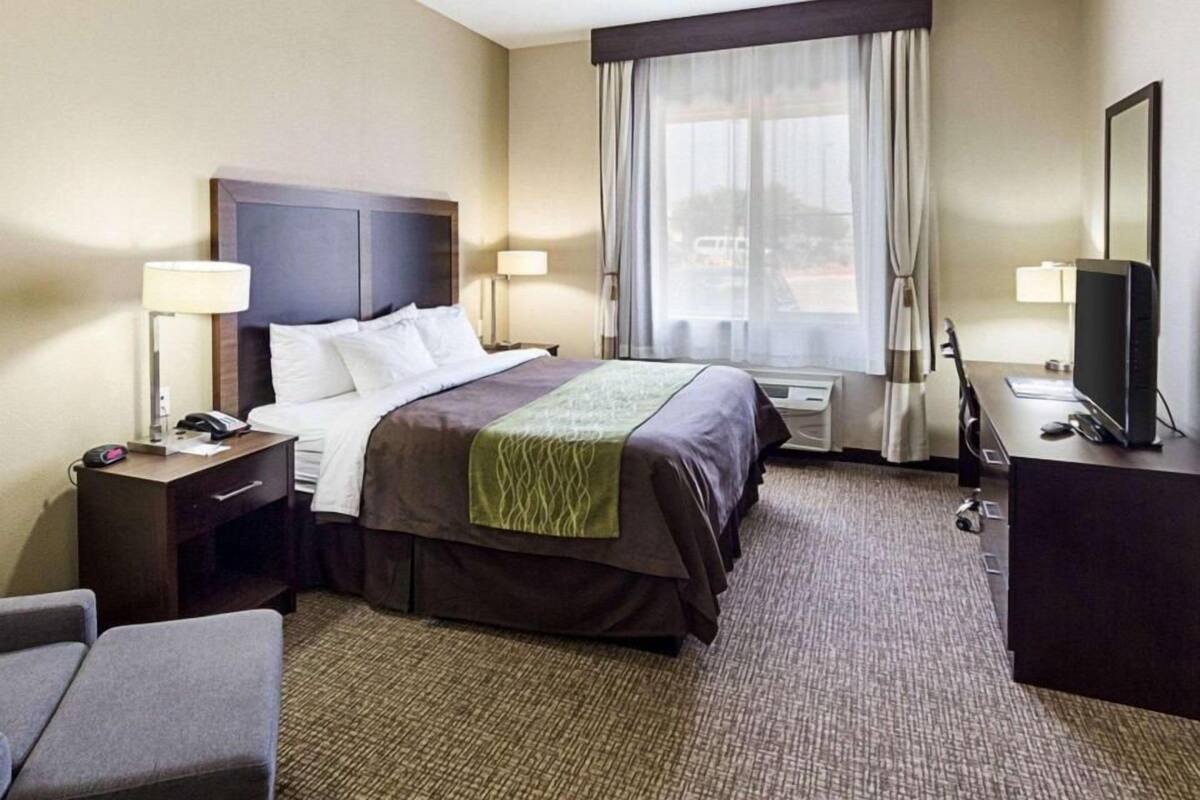 Quarto de hotel com uma cama, abajures, uma escrivaninha com computador, uma cadeira e uma janela com cortinas.
