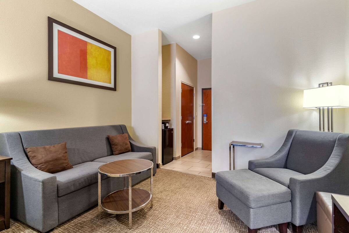 Quarto de hotel moderno com um sofá cinzento, uma mesa redonda pequena, uma poltrona cinzenta e uma luminária.