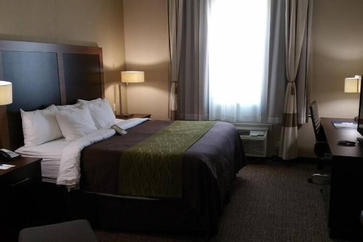 Quarto de hotel com uma cama, abajures, uma escrivaninha com computador e uma televisão.