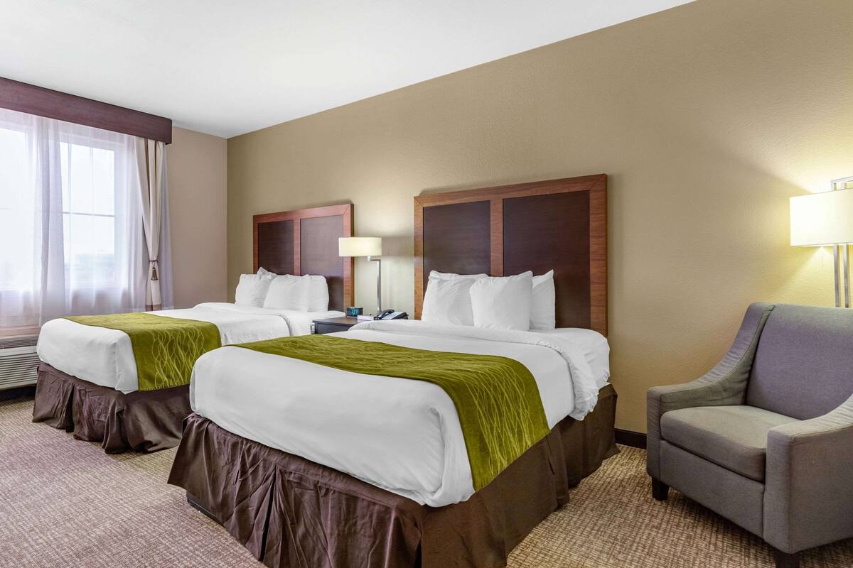Quarto de hotel com duas camas, uma cadeira, um abajur e uma janela com cortinas.
