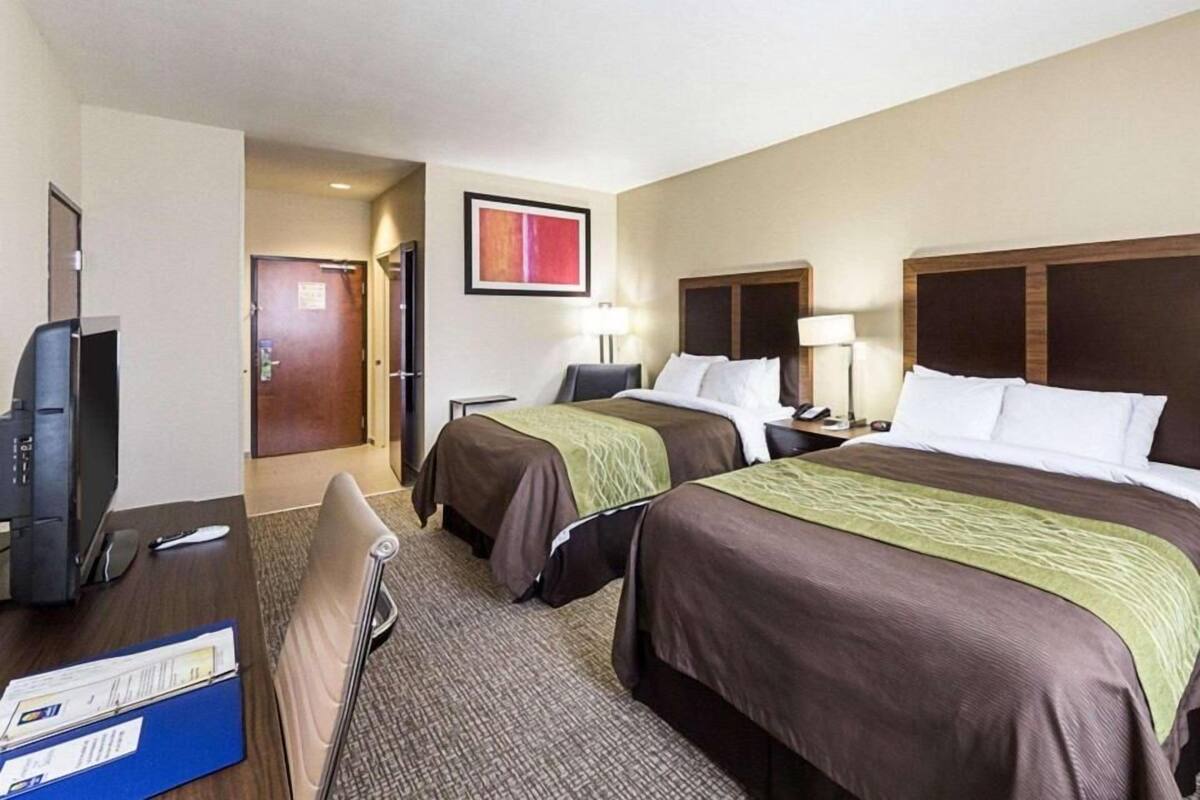 Quarto de hotel com duas camas, uma escrivaninha com computador, televisão e porta que dá para outro espaço.