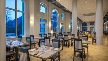 Uma sala de jantar ampla, com várias mesas e cadeiras, janelas grandes e um padrão de piso em espinha de peixe.