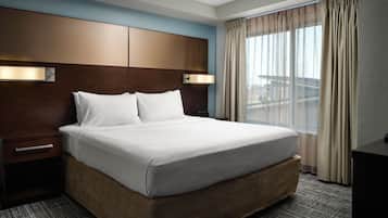 Quarto de hotel com uma cama grande, mesa de cabeceira, luminária na parede e janela com cortinas.
