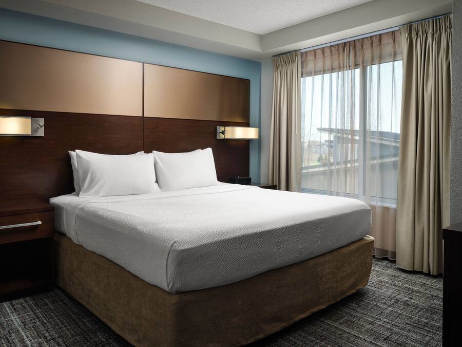 Quarto de hotel com uma cama grande, mesa de cabeceira, luminária na parede e janela com cortinas.