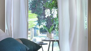 Quarto com cama, uma pequena mesa com xícara e pires, e um vaso com plantas de flores brancas.
