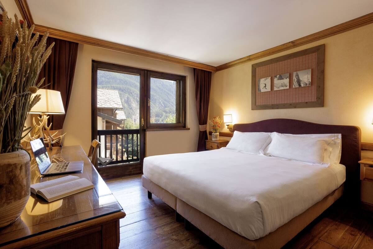 Quarto de hotel com uma cama, uma escrivaninha com um laptop, janela com vista para montanhas e um quadro na parede.