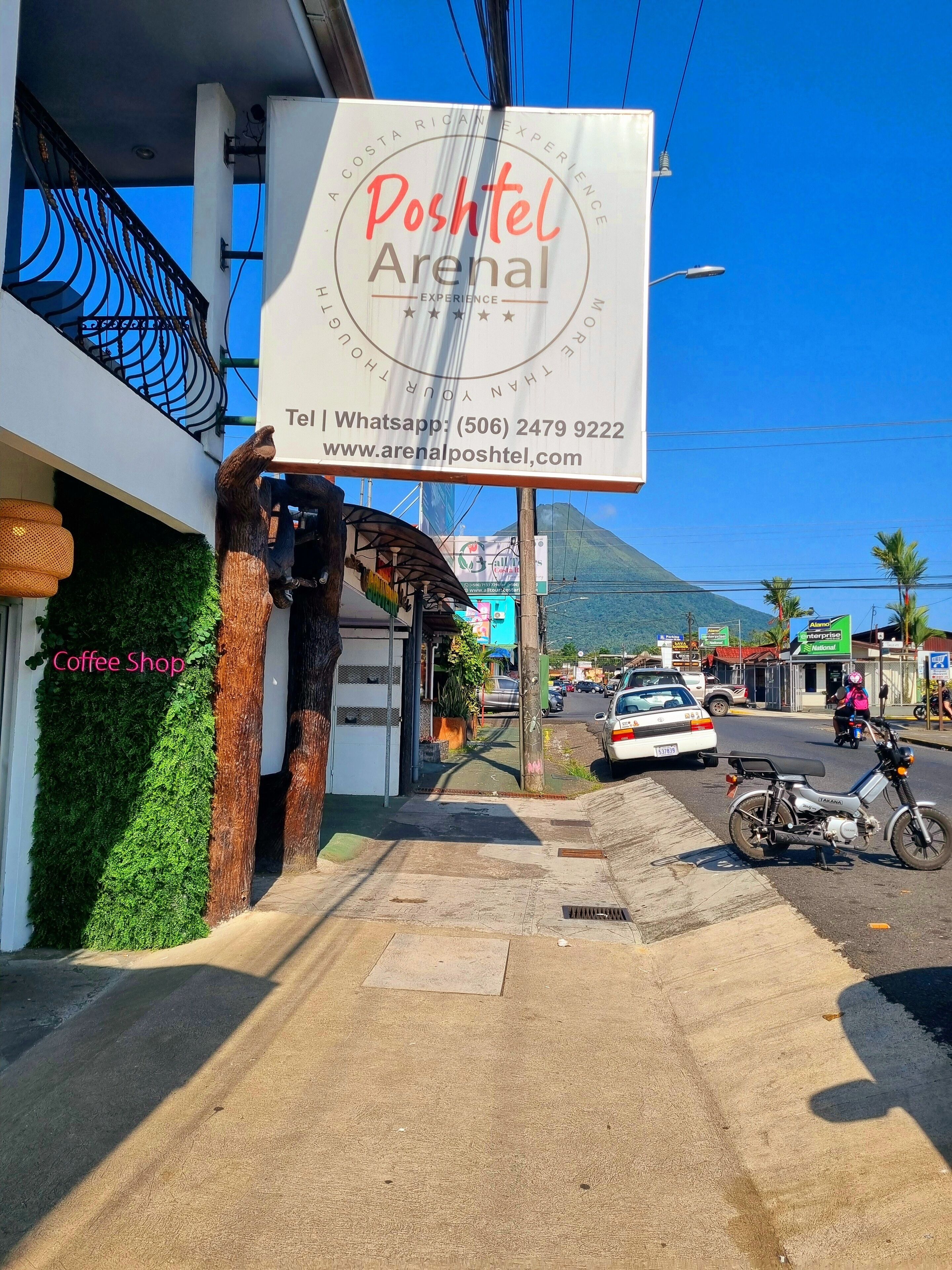 Vista da rua com a placa Poshtel Arenal, uma cafeteria e uma motocicleta estacionada.