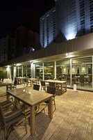Uma área de refeições ao ar livre com mesas e cadeiras, grandes janelas e um edifício ao fundo.