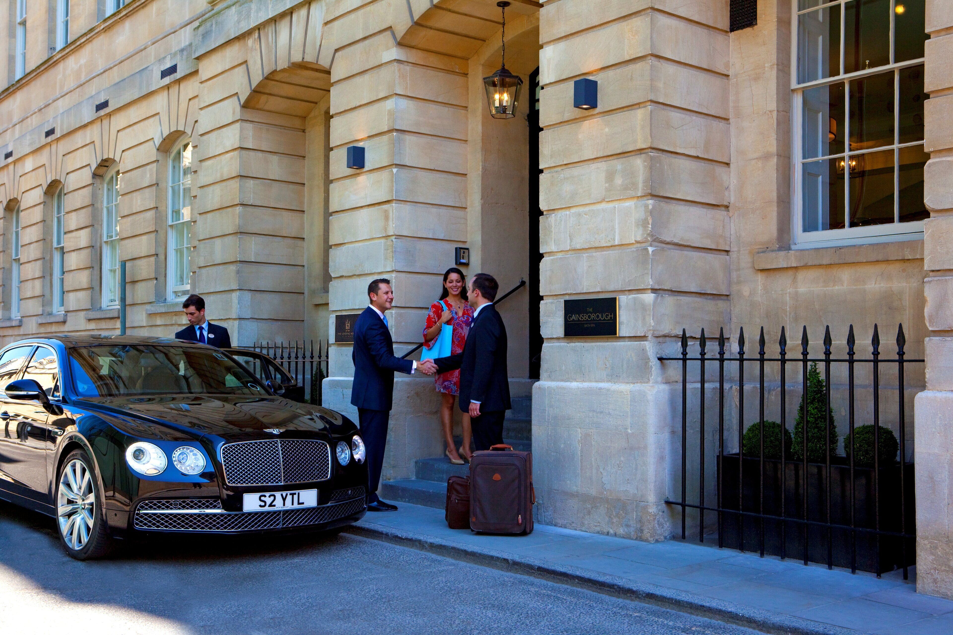 Um carro de luxo preto estacionado em frente a um edifício de arquitetura em pedra, uma cerca de metal preto e uma placa com a inscrição 'CAMBRIDGE HOSPITAL'.