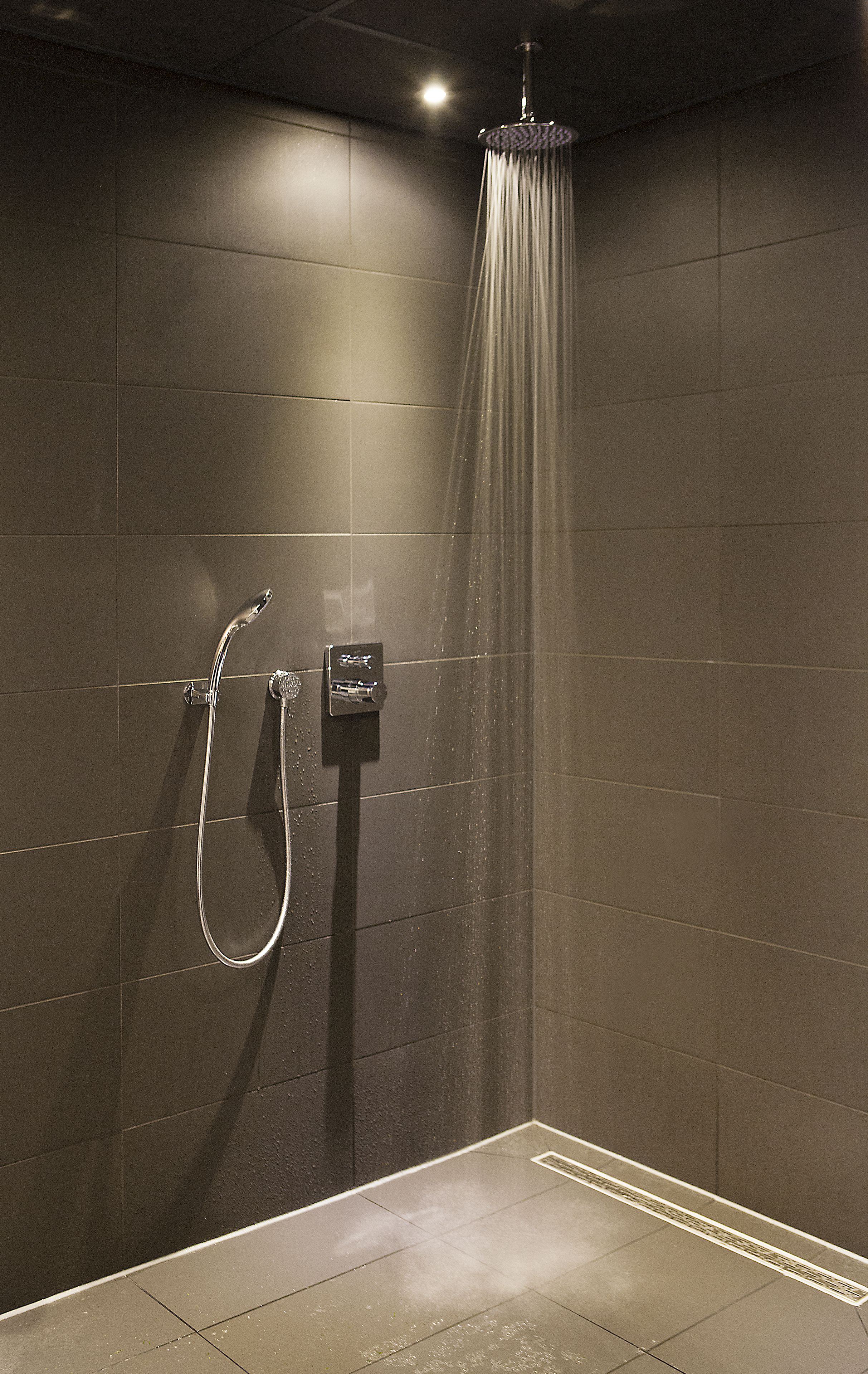 Área de banho moderna com chuveiro de efeito chuva e chuveiro manual, ambos instalados em parede com azulejos escuros.