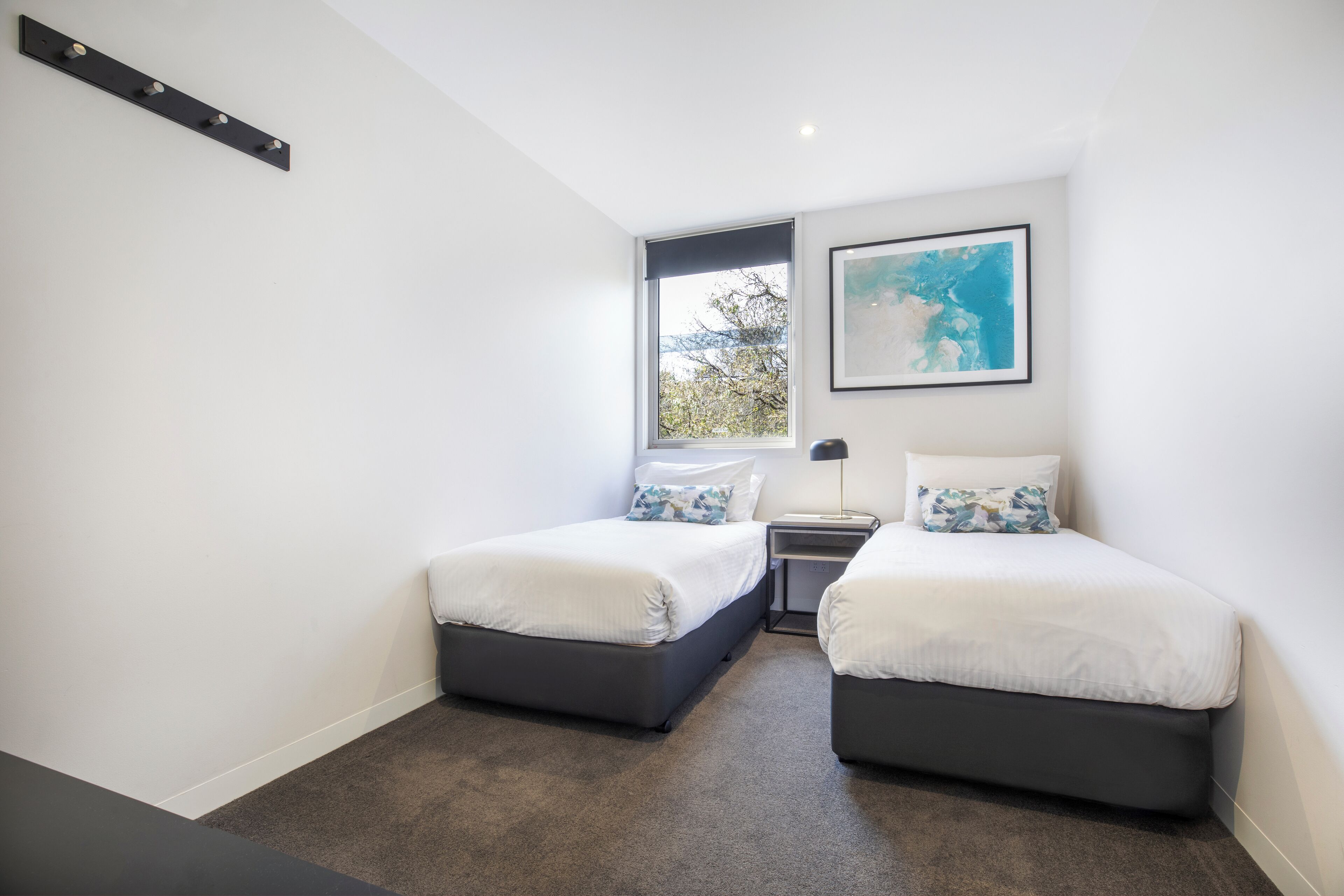 Quarto moderno com duas camas, uma janela, uma obra de arte emoldurada e uma mesa de cabeceira.