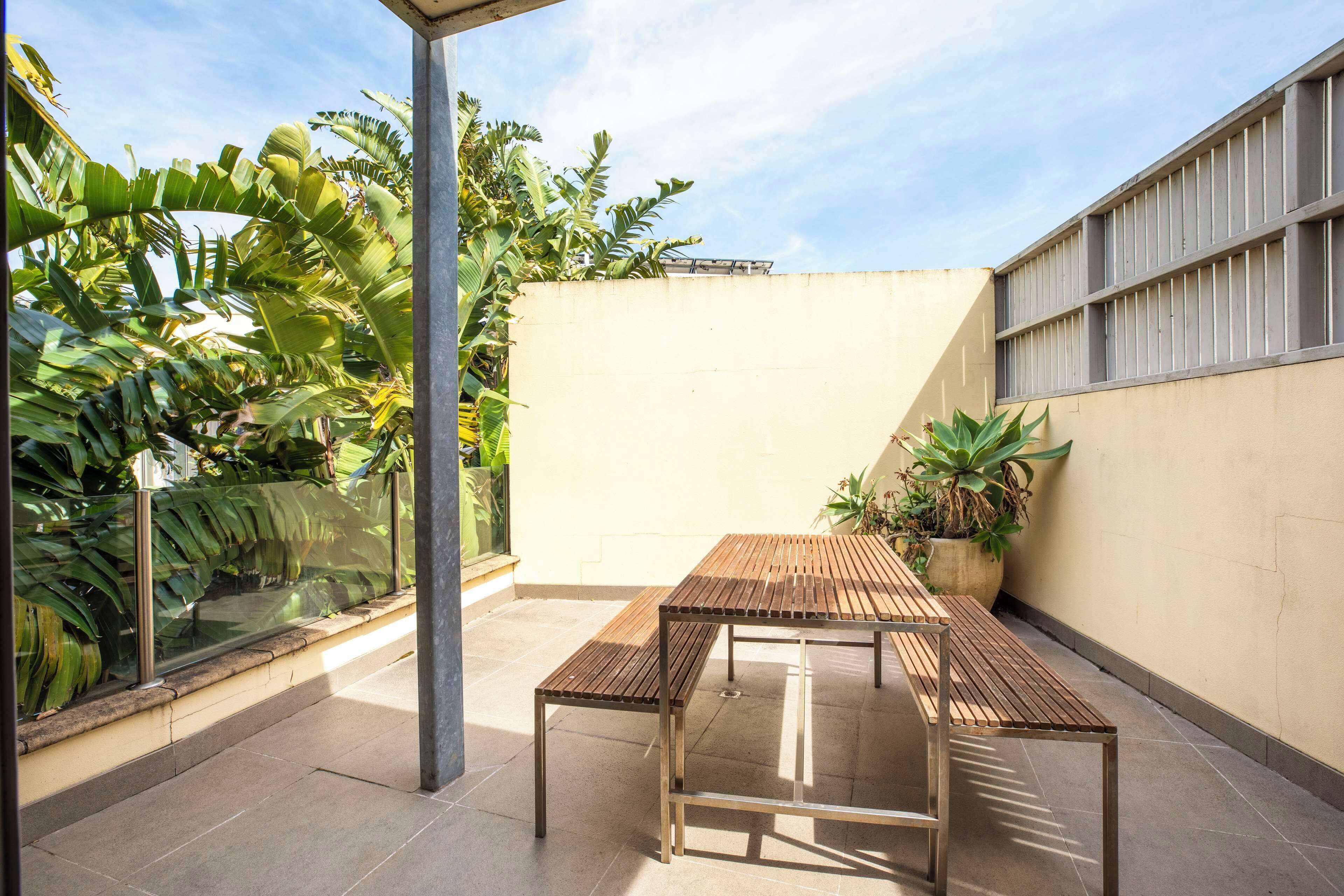 Um pátio externo com mesa e cadeiras de madeira, rodeado por plantas em vasos e um guarda-corpo de vidro.