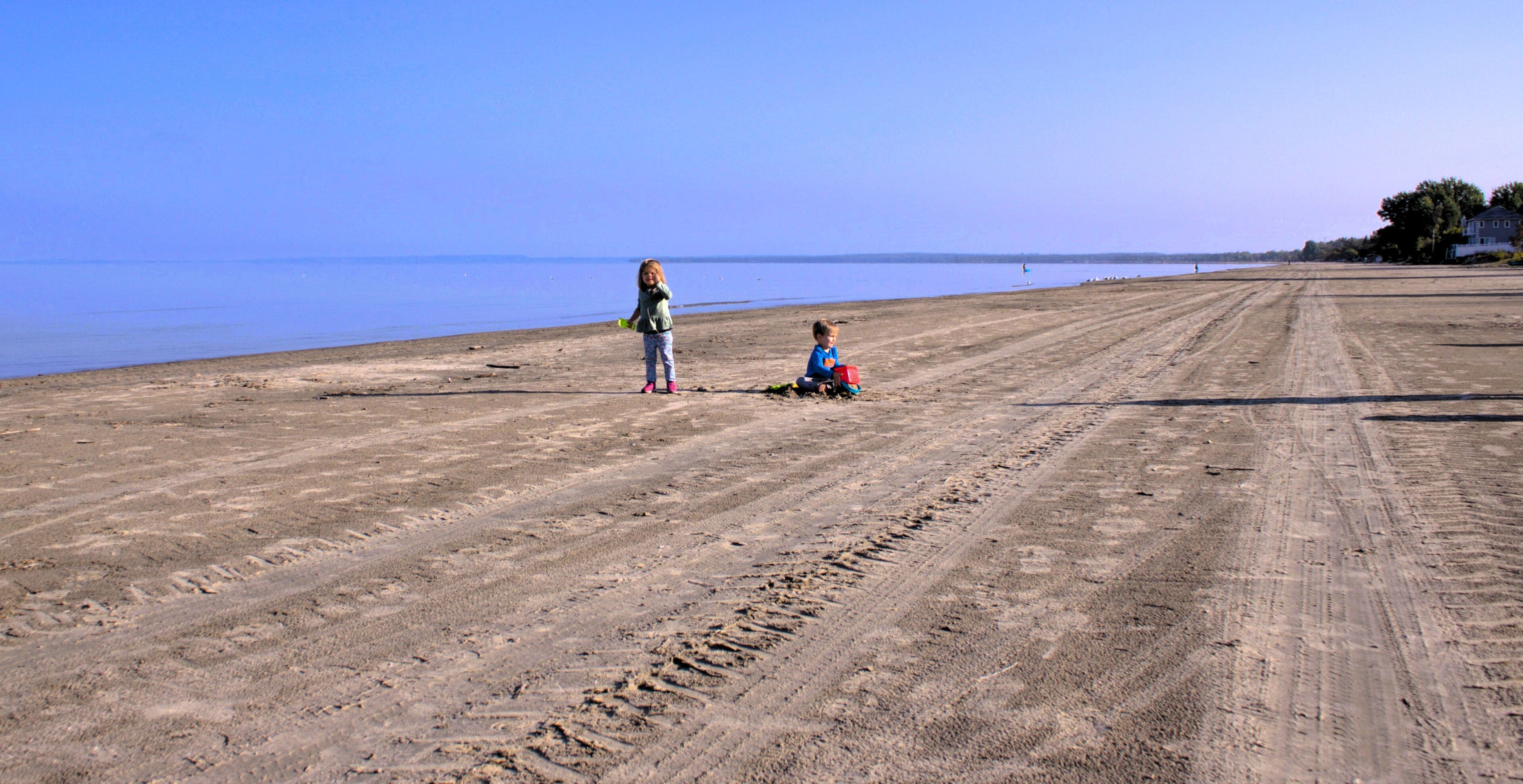 Uma praia arenosa com pegadas, uma criança brincando e uma pessoa parada ao longe.