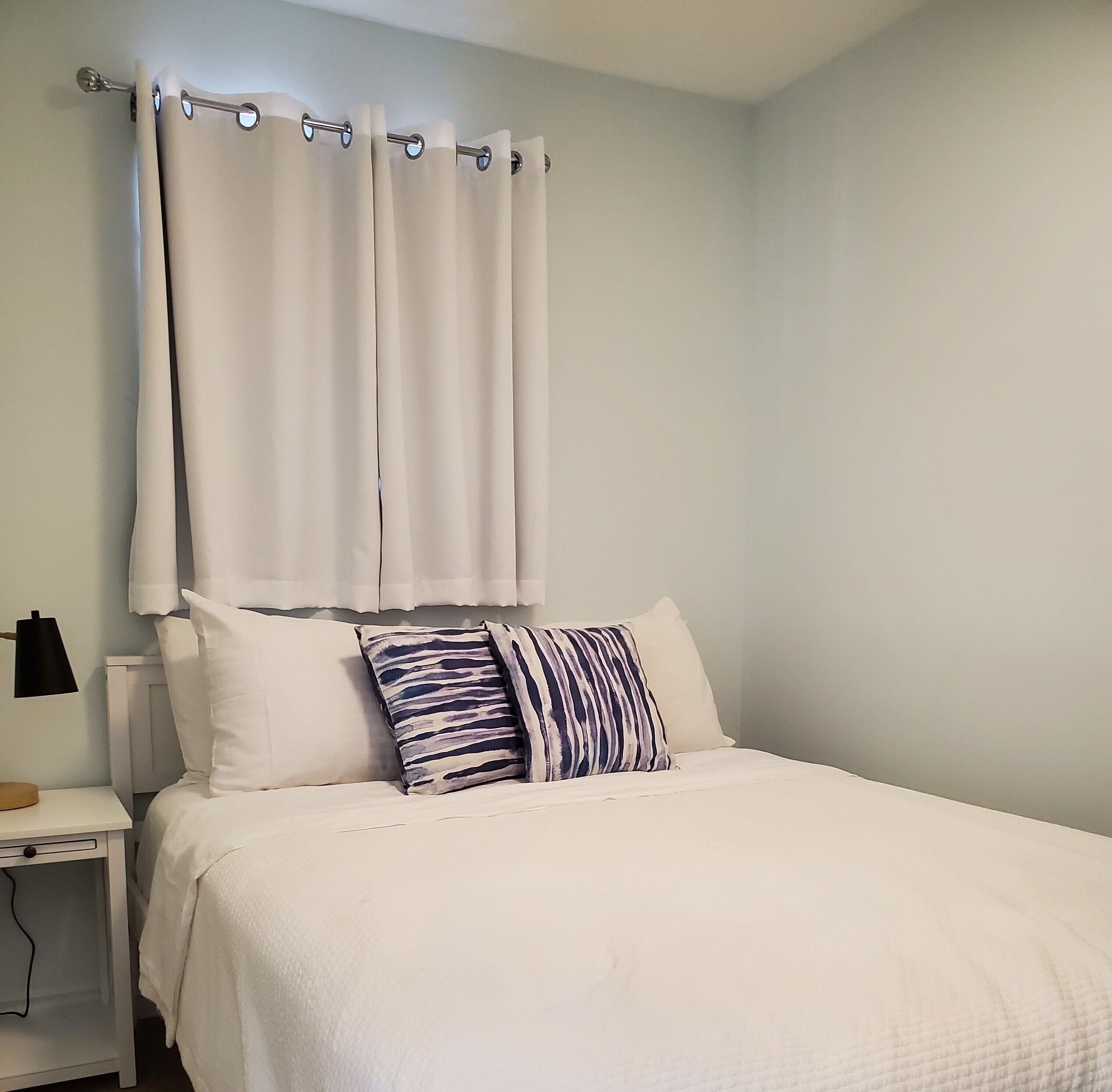 Quarto com cama, cortinas brancas, mesa de cabeceira com abajur e travesseiro estampado com listras.