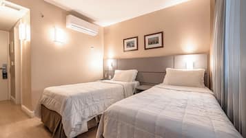 Quarto de hotel com duas camas de solteiro, mesa de cabeceira com abajur, aparelho de ar condicionado e quadros na parede.