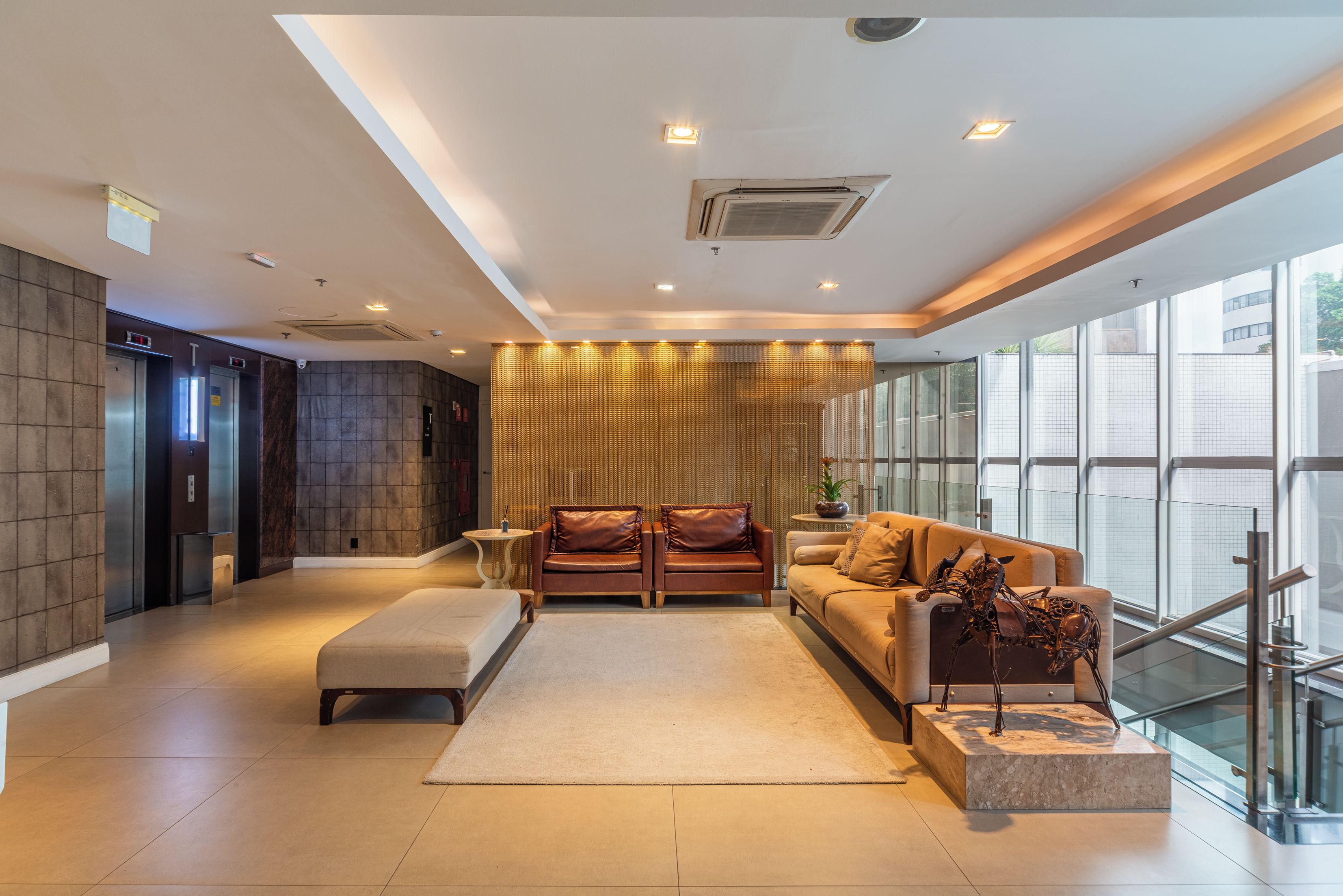 Um lobby moderno com área de estar, painel de elevador digital e uma grande janela com vista para o exterior.