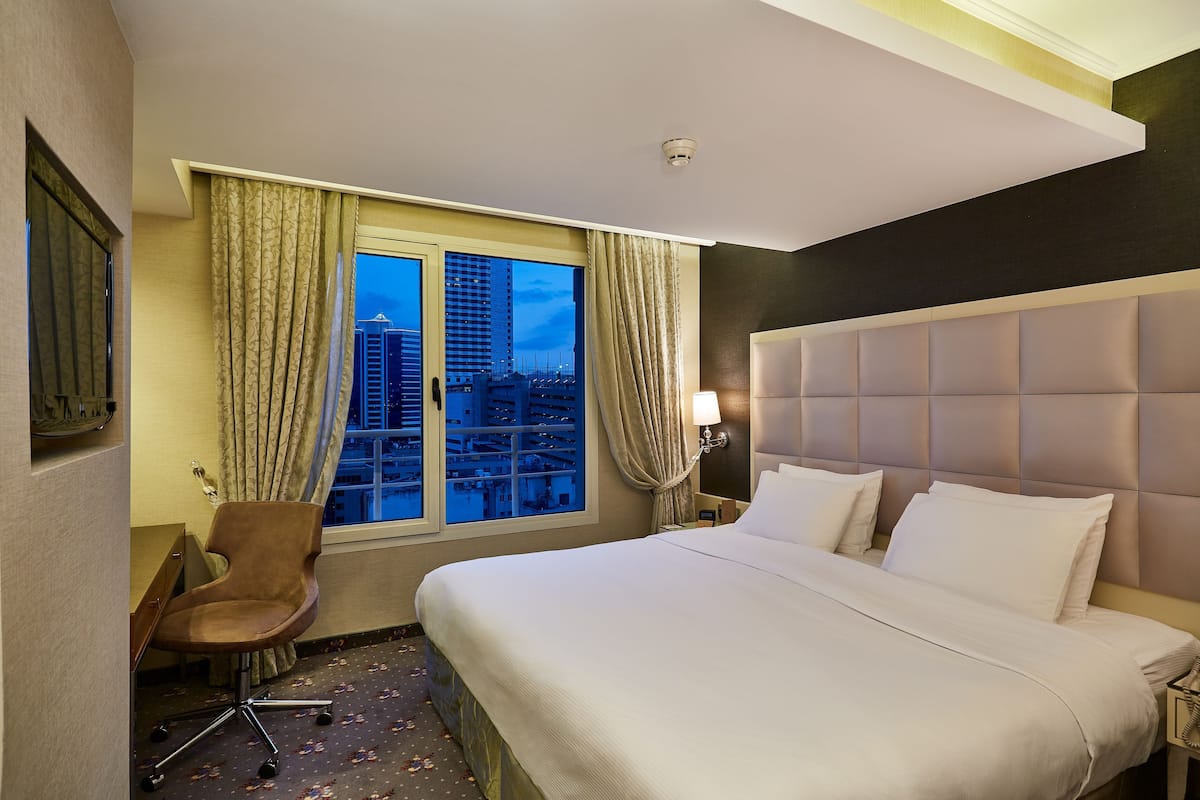 Quarto de hotel com cama, escrivaninha, cadeira, janela com cortinas e vista para a cidade.