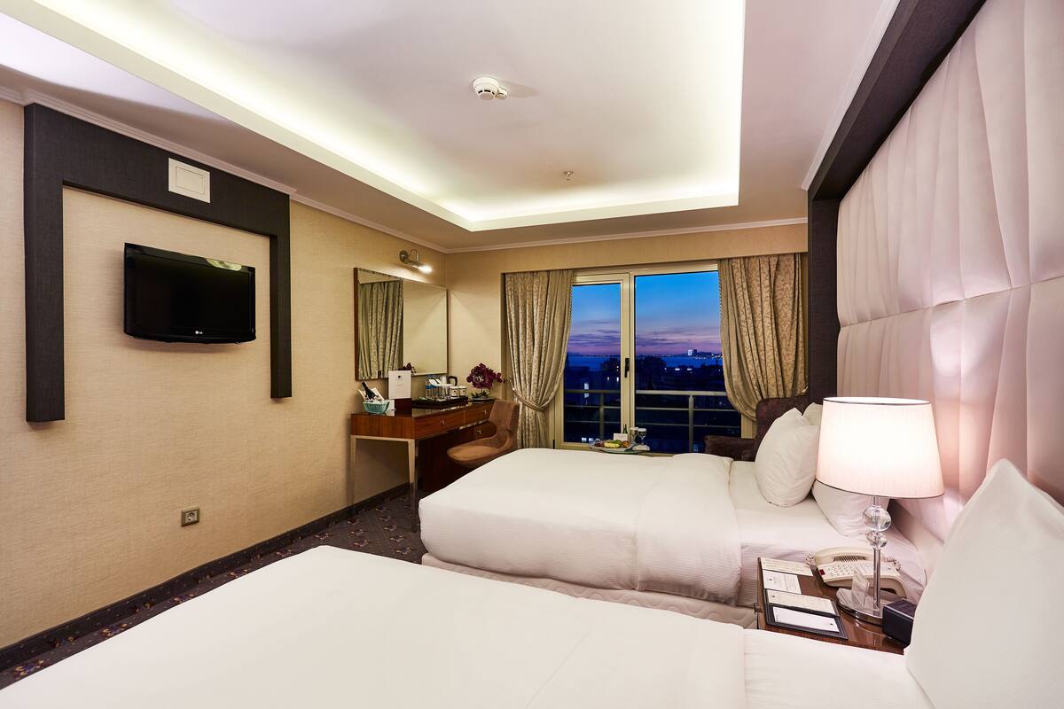 Quarto de hotel com duas camas, uma escrivaninha, televisão e janela com vista para a cidade.