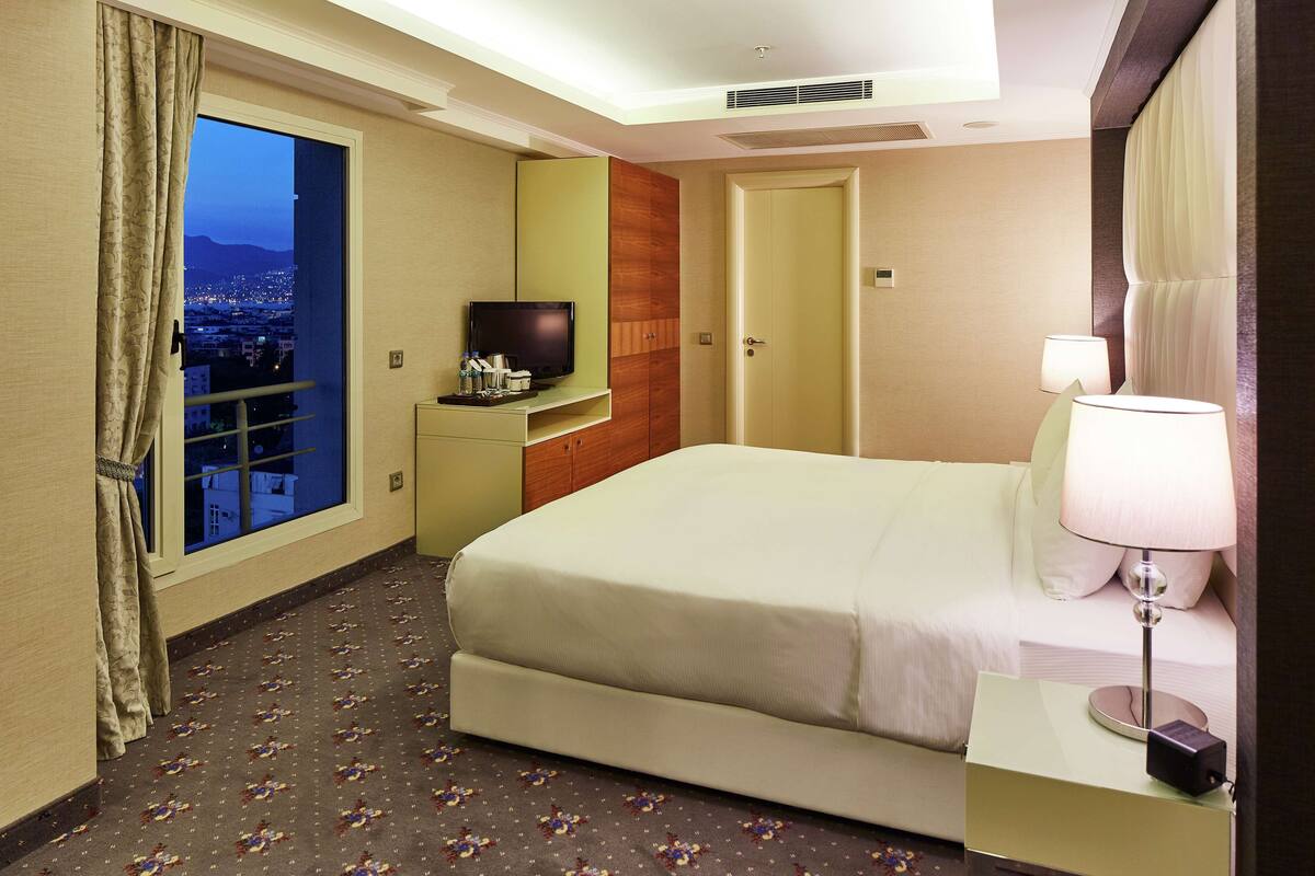 Quarto de hotel com uma cama, televisão, mesa de cabeceira, abajur e vista para a cidade através da janela.