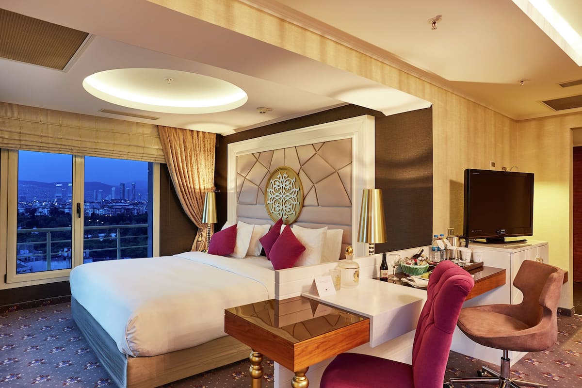 Quarto de hotel com uma cama grande, uma mesa com cadeira, televisão e vista da cidade pela janela.