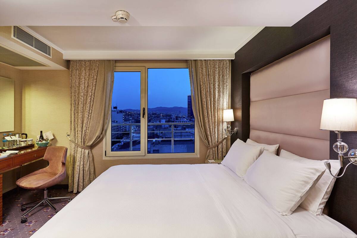 Quarto de hotel com uma cama grande, uma escrivaninha com cadeira, uma luminária de mesa e vista para a cidade pela janela.