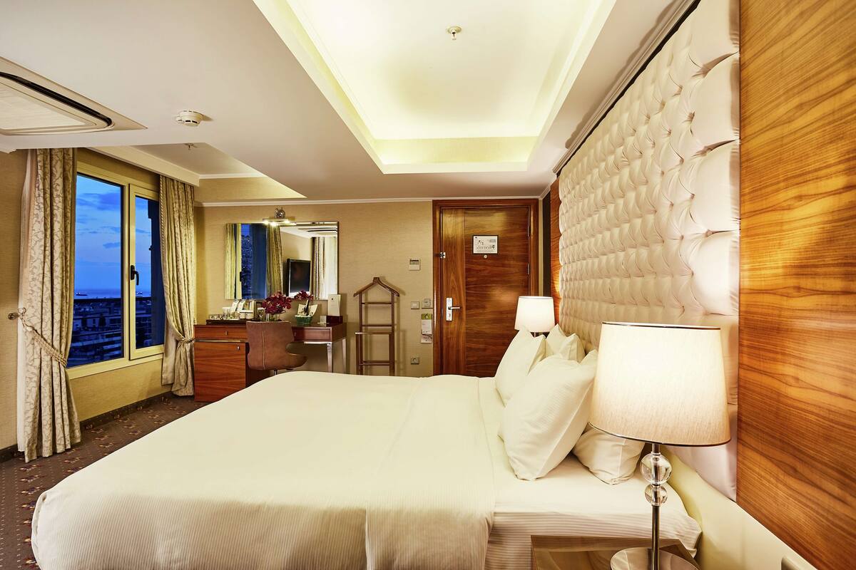 Quarto de hotel com uma cama grande, uma luminária de cabeceira, cabeceira de madeira, cômoda, espelho e vista da cidade por uma janela ampla.