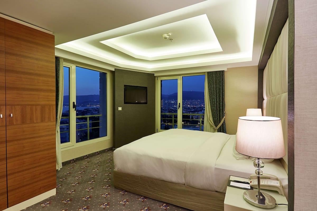 Quarto de hotel com uma cama grande, uma mesa com luminária, telefone, TV e vista da cidade através de amplas janelas.