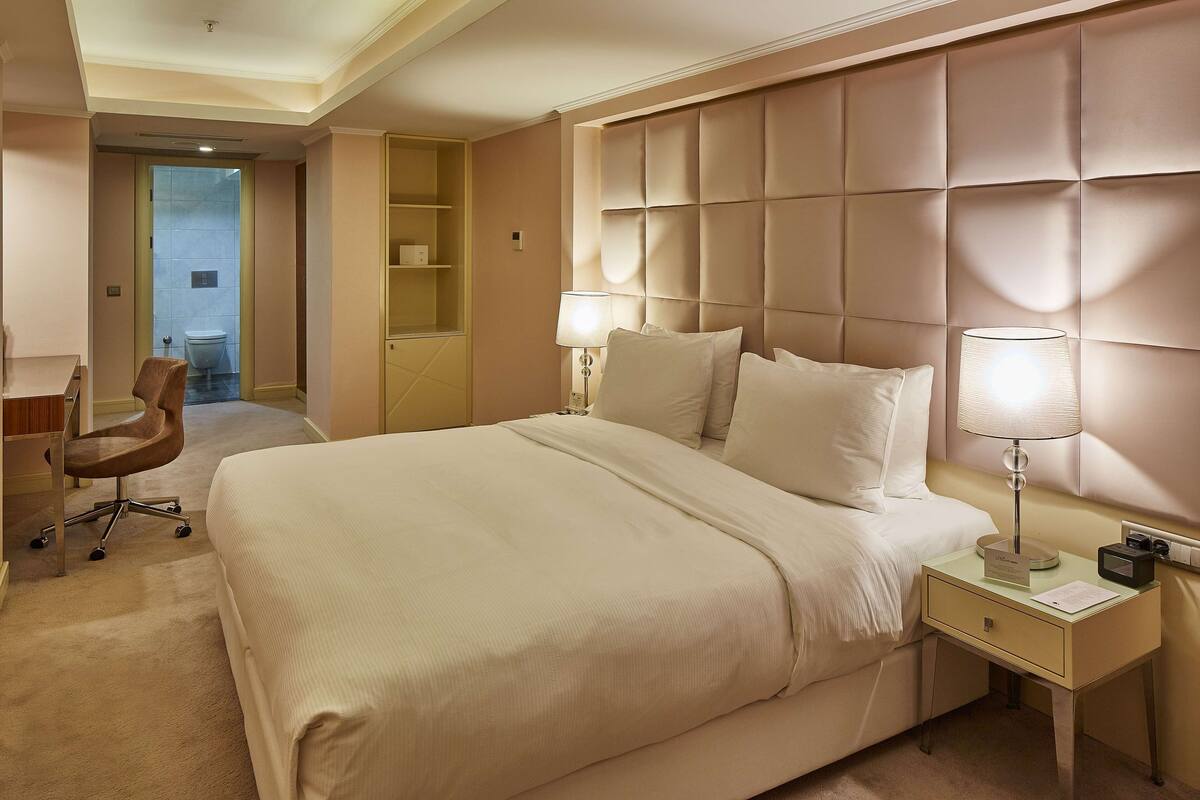 Quarto de hotel com uma cama grande, uma escrivaninha, uma cadeira e uma mesa de cabeceira com abajur.
