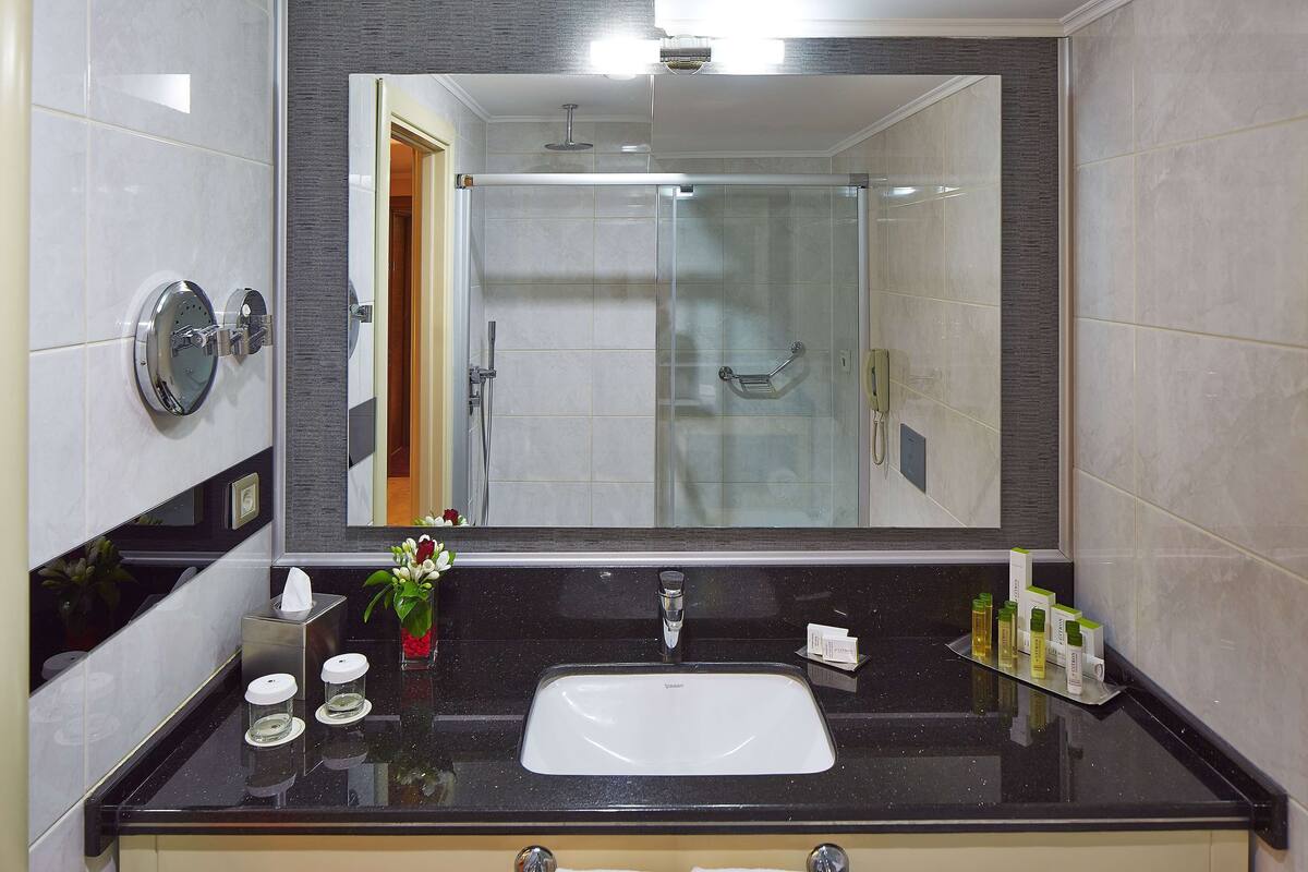 Casa de banho com bancada preta, lavatório branco, cabine de duche com vidro e um espelho grande.