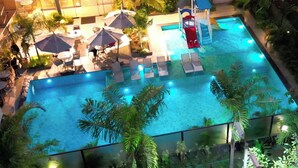 Piscina com espreguiçadeiras e guarda-sóis, cercada por palmeiras e um edifício com uma placa.