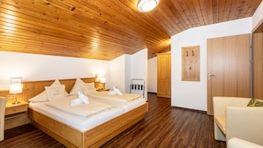 Quarto de hotel com teto de madeira, uma cama grande com roupa de cama branca, mesa de cabeceira com luminária e guarda-roupa de madeira.