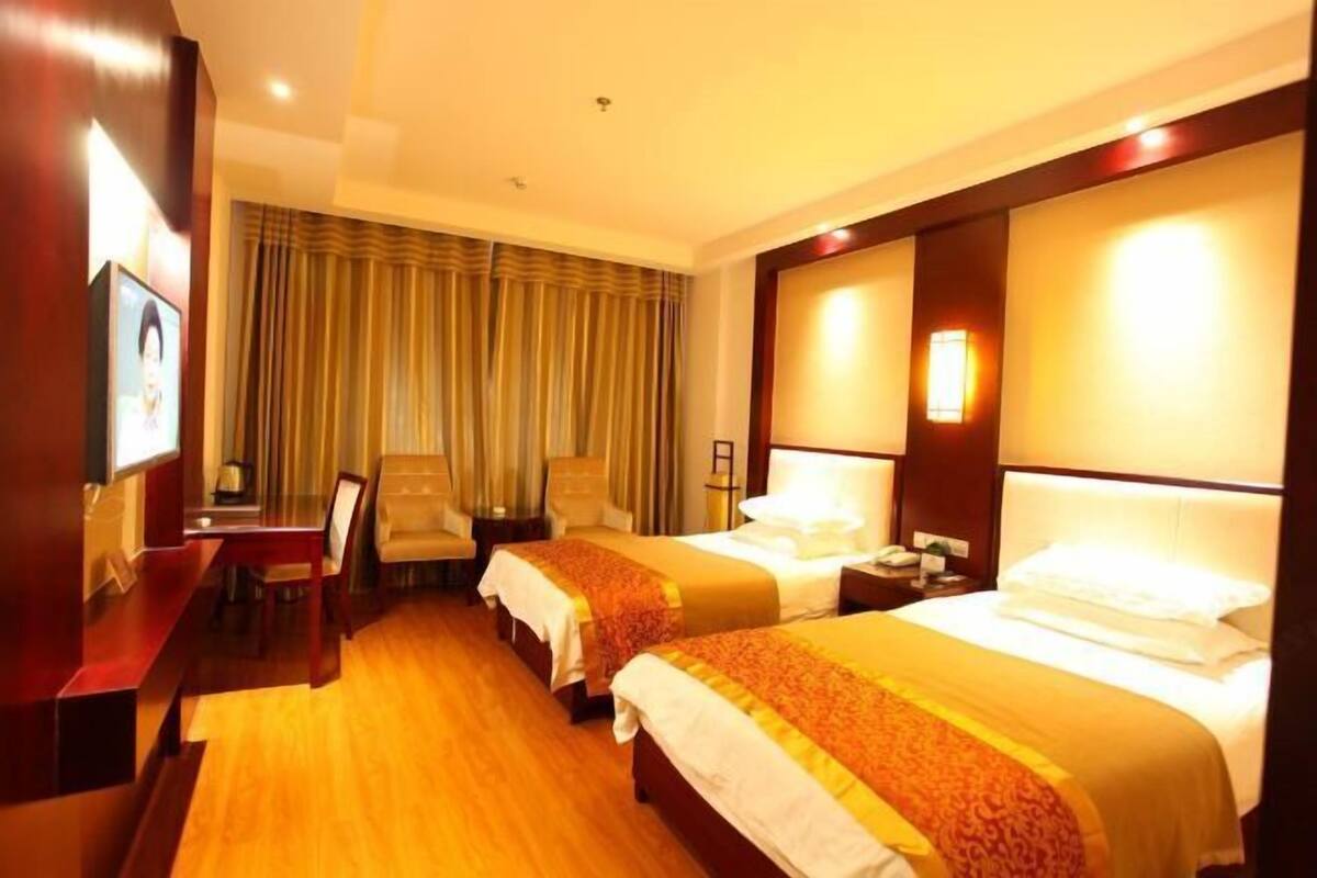 Quarto de hotel com duas camas, uma escrivaninha, uma cadeira, televisão e uma janela grande com cortinas.