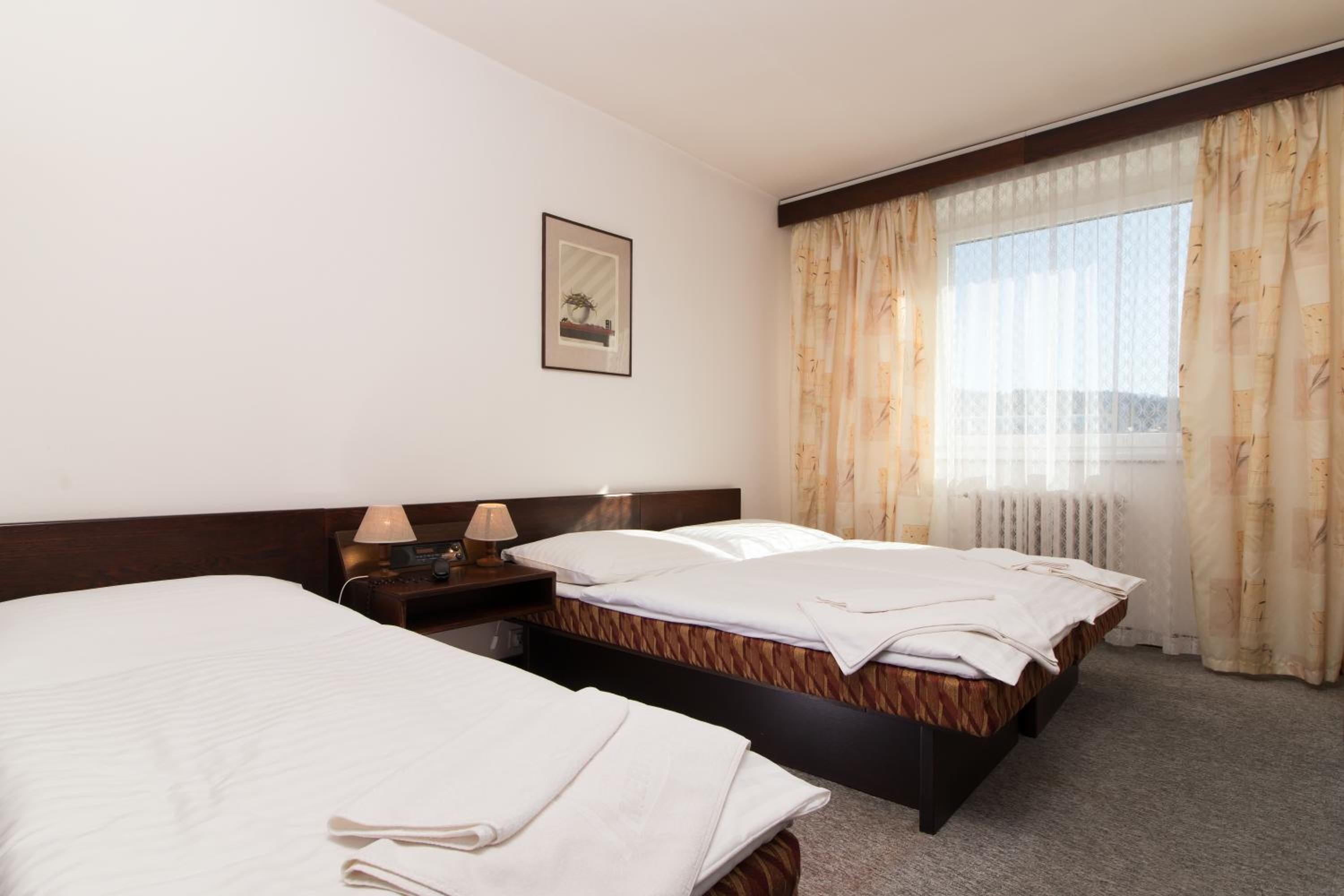 Quarto de hotel com duas camas, uma mesinha de cabeceira com luminárias, um quadro na parede e uma janela com cortinas.