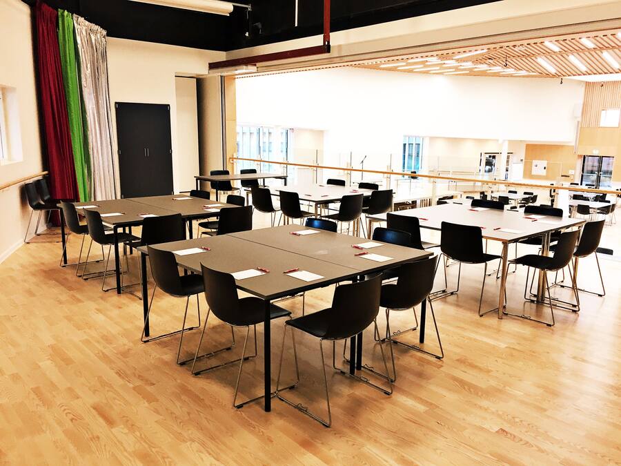 Sala de conferências com várias mesas e cadeiras, piso de madeira e amplas janelas que permitem a entrada de luz natural.