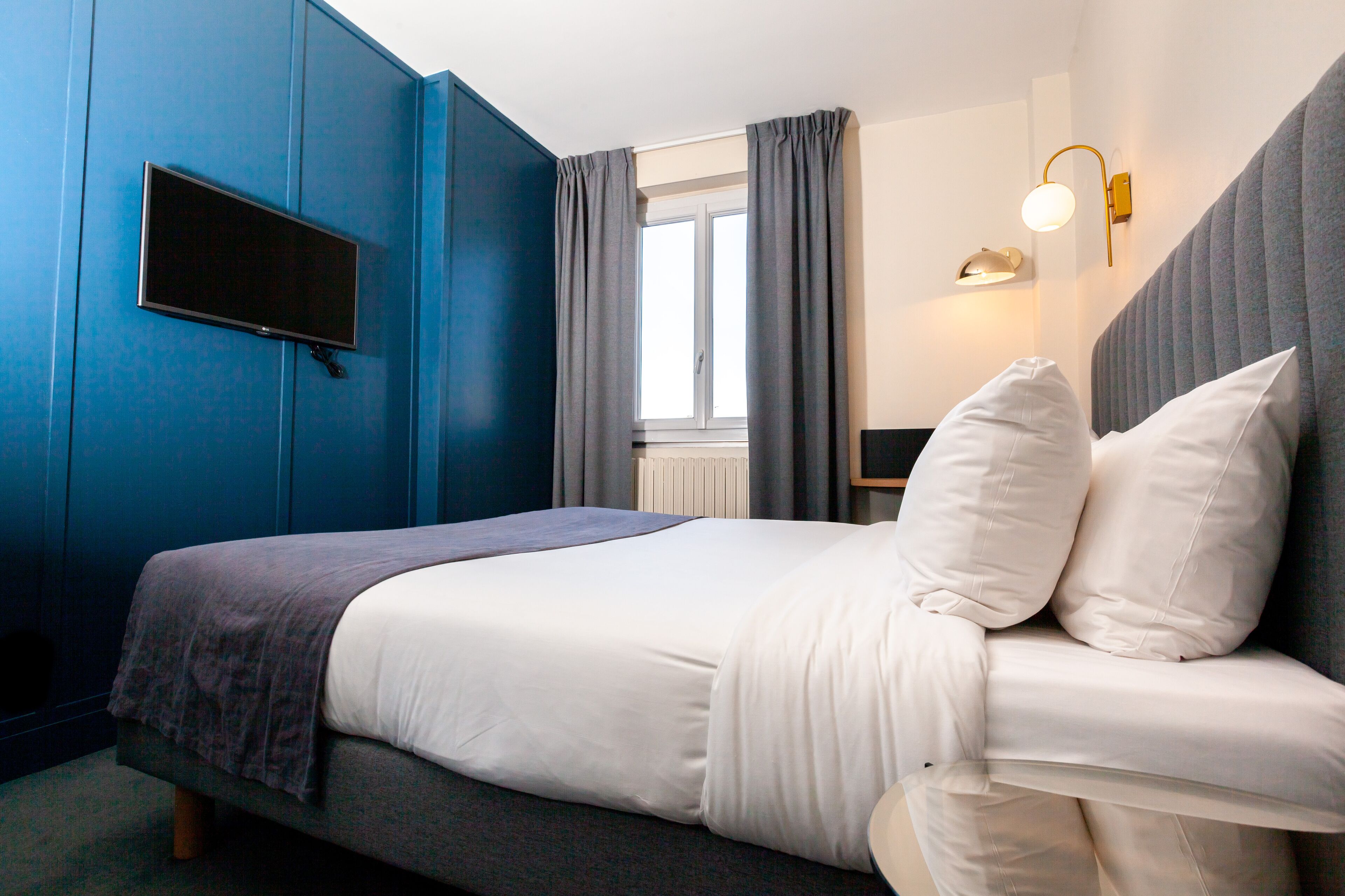 Quarto de hotel com cama, televisão de tela plana na parede, janela com cortinas e abajur.
