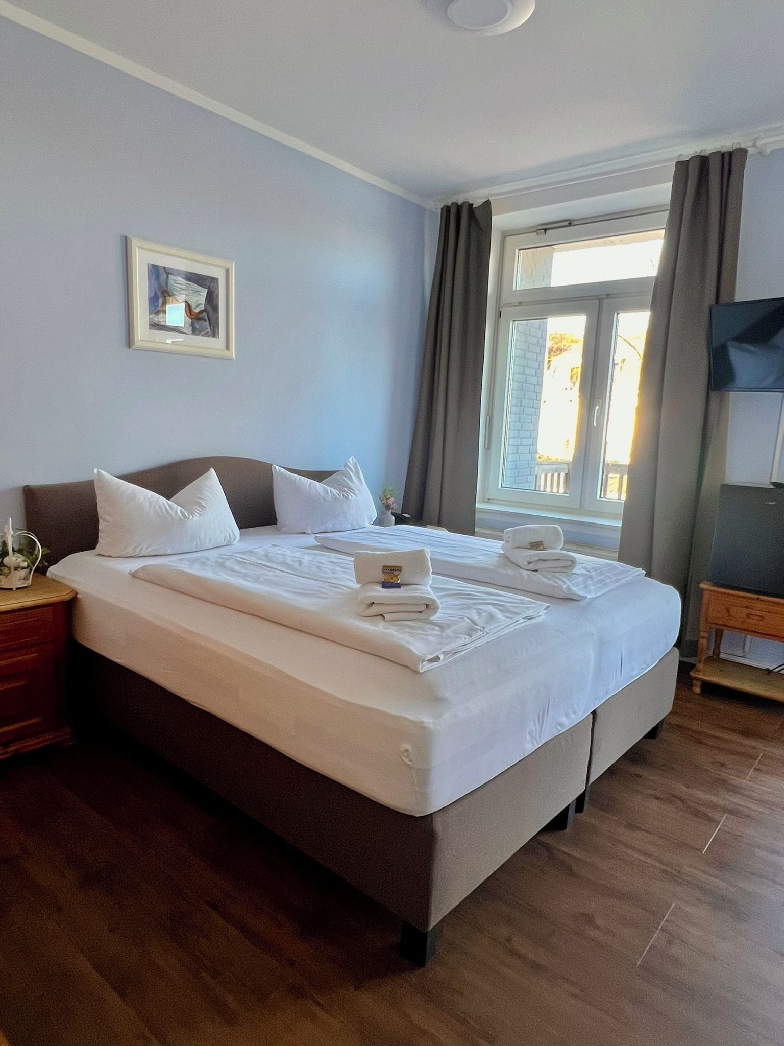 Quarto de hotel com cama grande, mesa de cabeceira, televisão e janela com cortinas.
