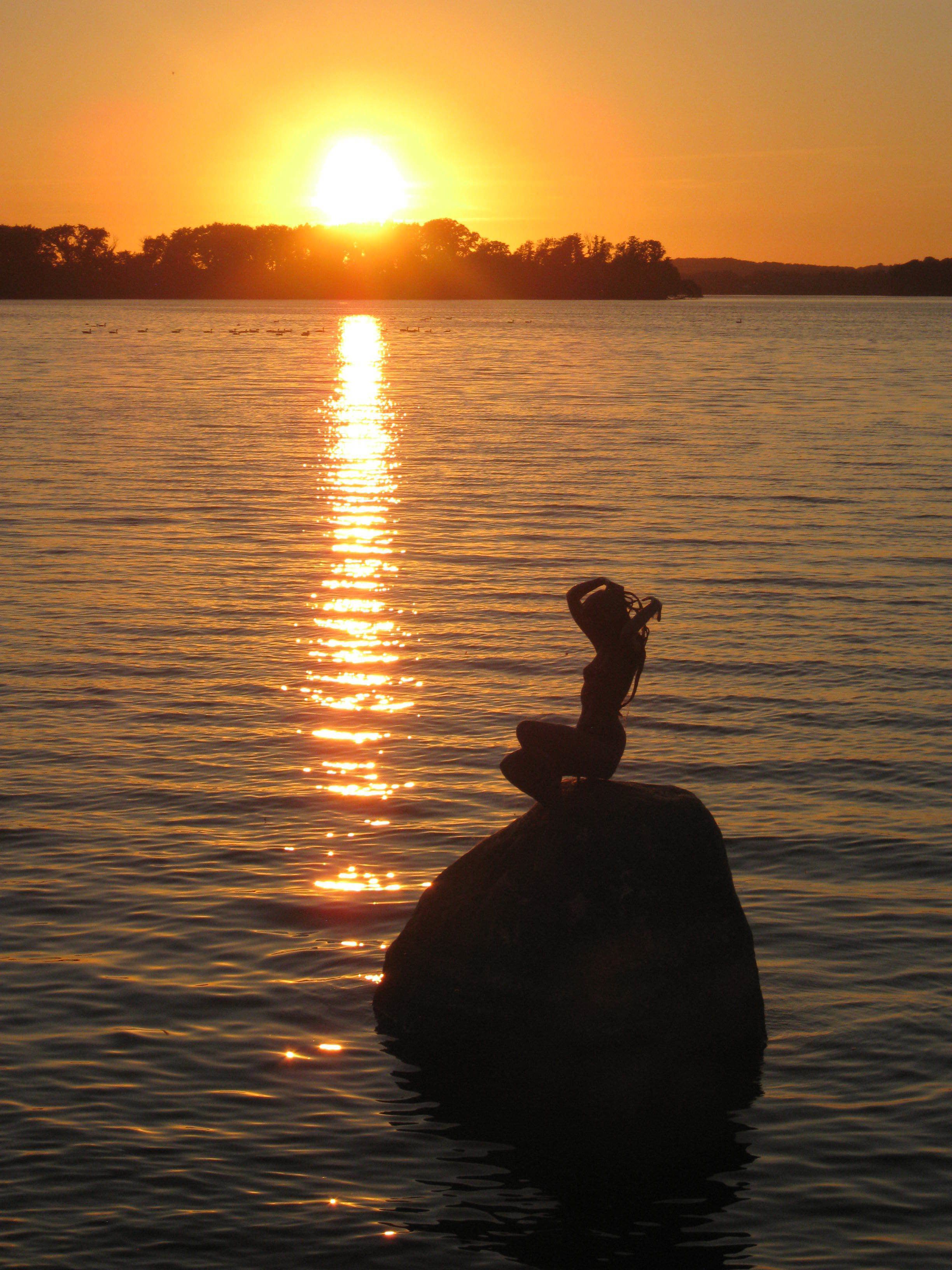Um pôr do sol sobre a água, com a silhueta de uma pessoa numa rocha.