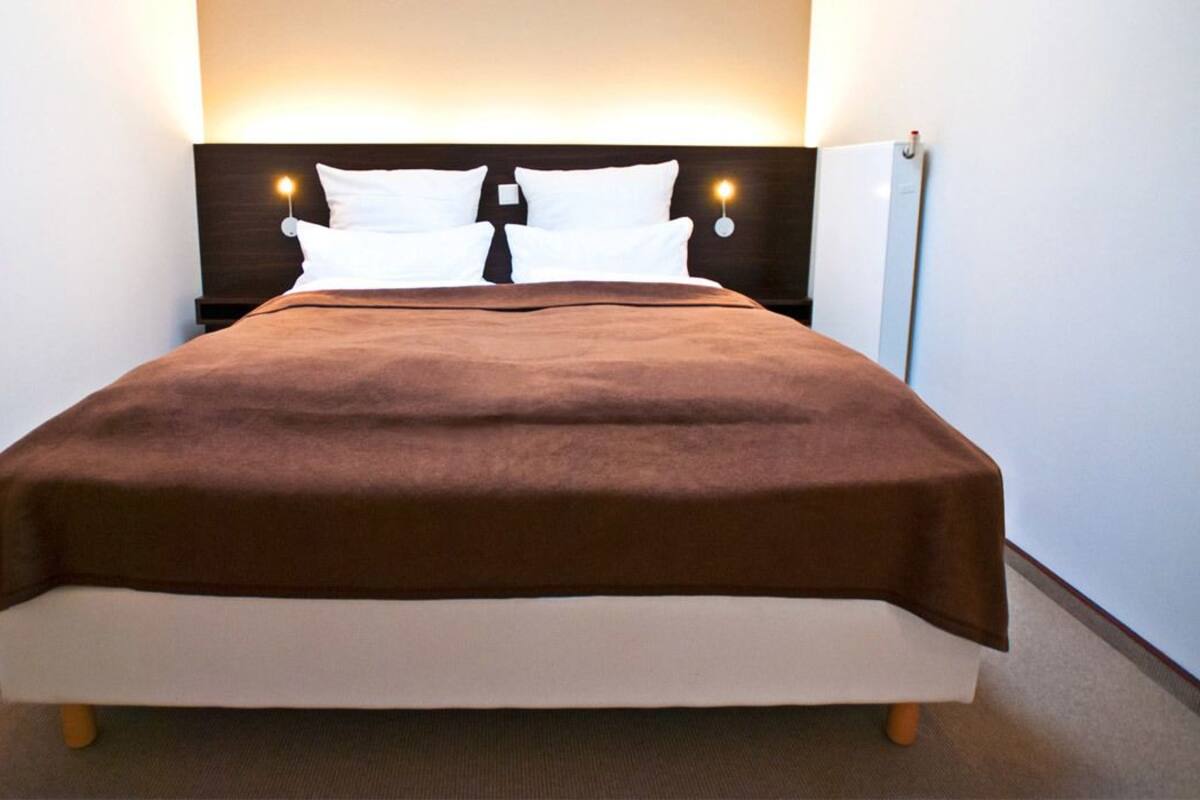 Cama bem arrumada com colcha marrom e almofadas brancas, cabeceira escura com luminárias fixadas na parede e mesa de cabeceira branca.