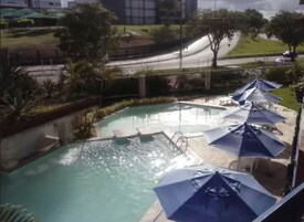 Piscina com vários guarda-sóis e espreguiçadeiras, rodeada por vegetação e uma estrada ao fundo.
