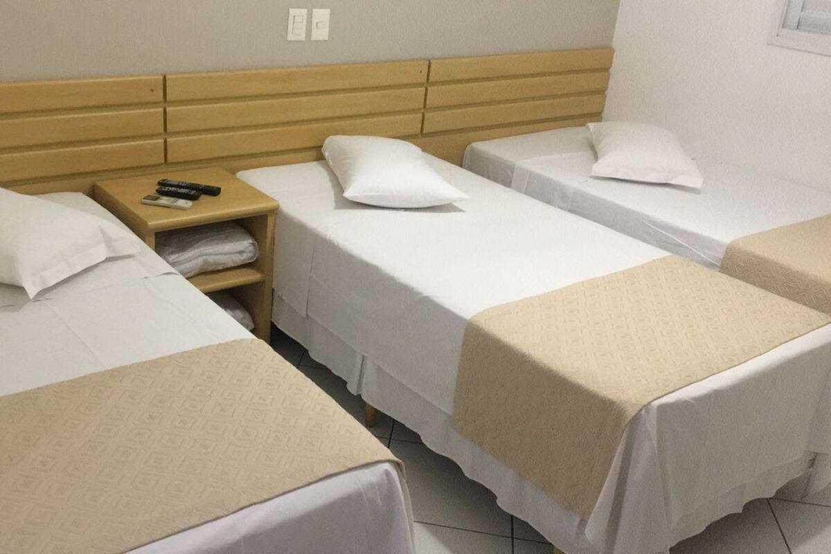 Quarto de hotel com três camas de solteiro, uma mesinha de cabeceira de madeira com uma toalha e uma pequena mesa.