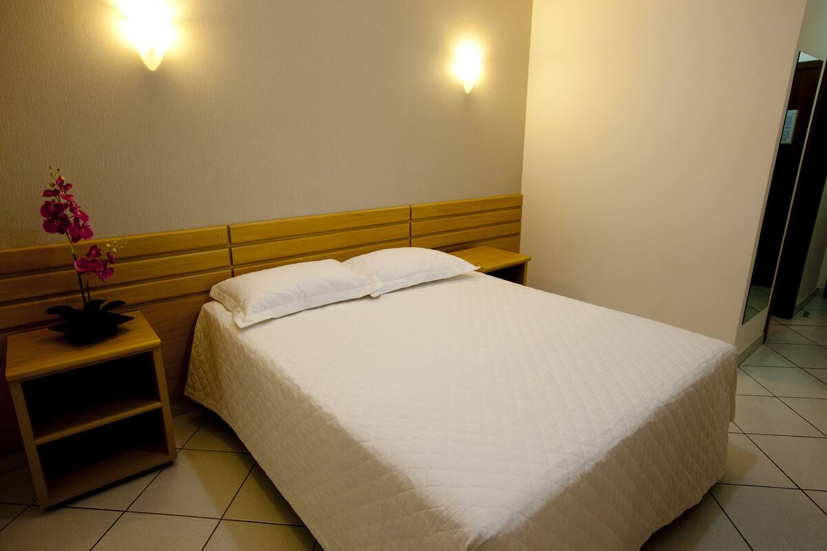 Uma cama bem arrumada com cabeceira de madeira, duas mesas de cabeceira e um vaso com uma flor.