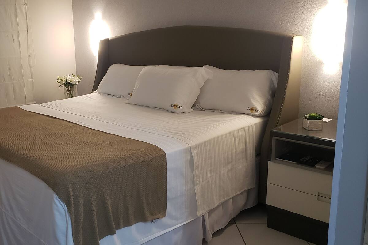 Cama bem arrumada com manta marrom, mesa de cabeceira com abajur e uma pequena planta.